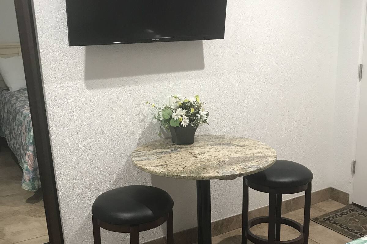 Um quarto pequeno com uma mesa redonda, dois banquinhos, um vaso de planta, uma TV de tela plana e uma cama com colcha estampada.