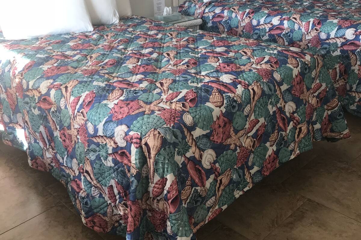 Quarto de hotel com duas camas, cada uma com almofadas brancas e edredom estampado com motivos florais.