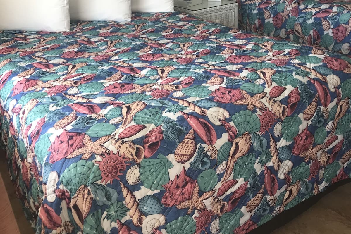 Uma cama de casal com colcha estampada em flor, duas almofadas brancas e uma mesa de cabeceira com abajur.