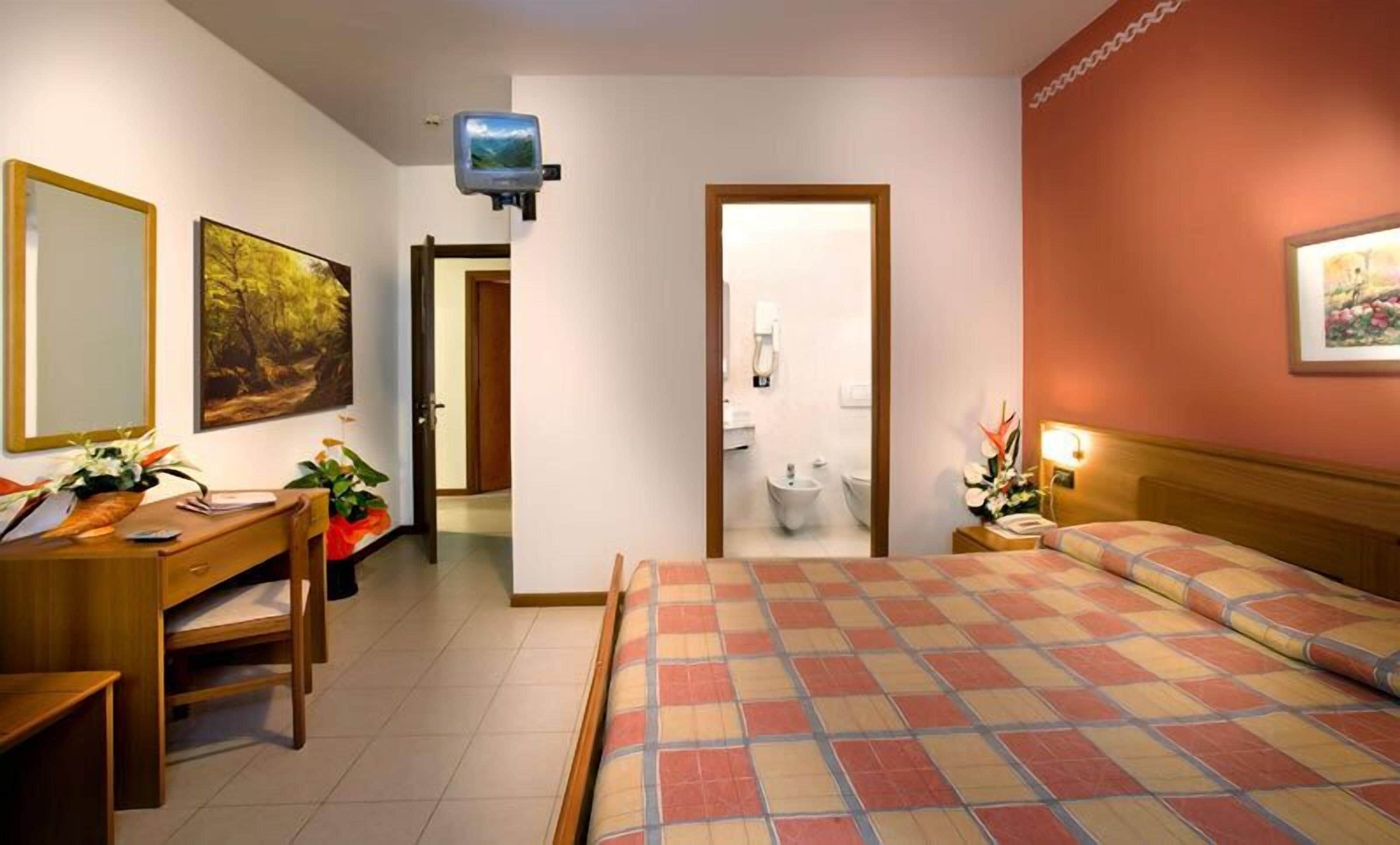 Quarto de hotel com cama, escrivaninha, cadeira, televisão na parede e banheiro visível por uma porta aberta.