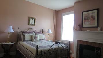 Quarto com cama de dossel, duas luminárias de cabeceira, um quadro na parede, lareira e janela com cortinas.