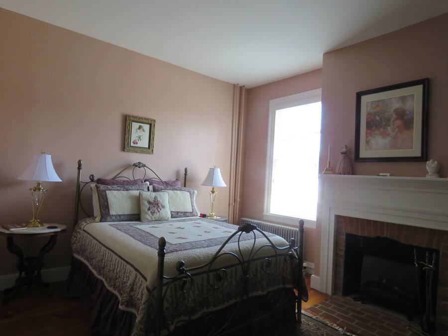 Quarto com cama de dossel, duas luminárias de cabeceira, um quadro na parede, lareira e janela com cortinas.
