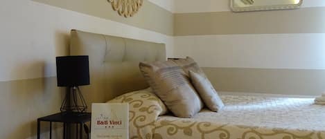 Quarto de hotel com cama, mesa de cabeceira com luminária, um objeto decorativo na parede e espelho emoldurado.