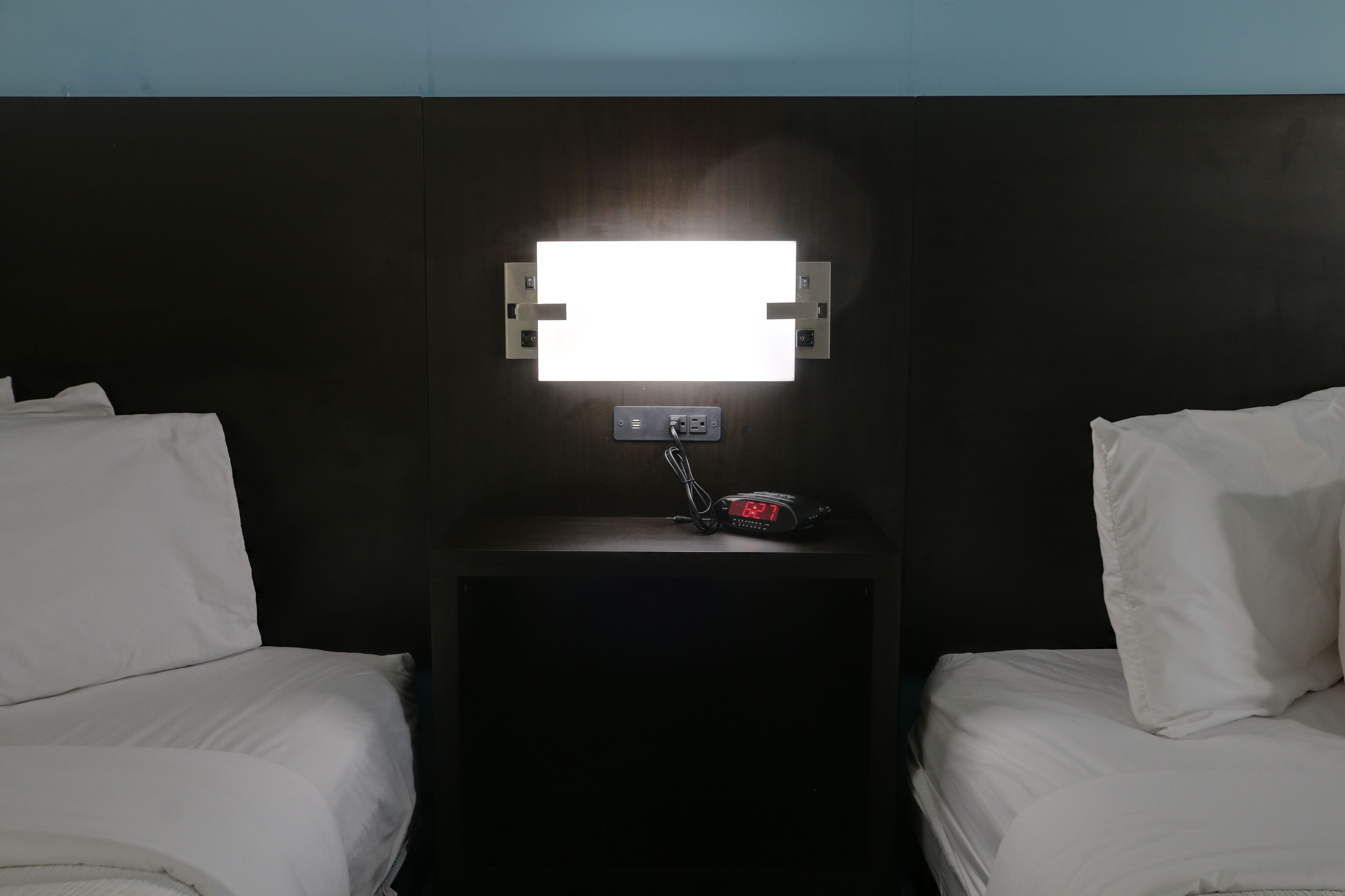 Quarto de hotel com duas camas, uma mesinha de cabeceira com abajur e luminária na parede.