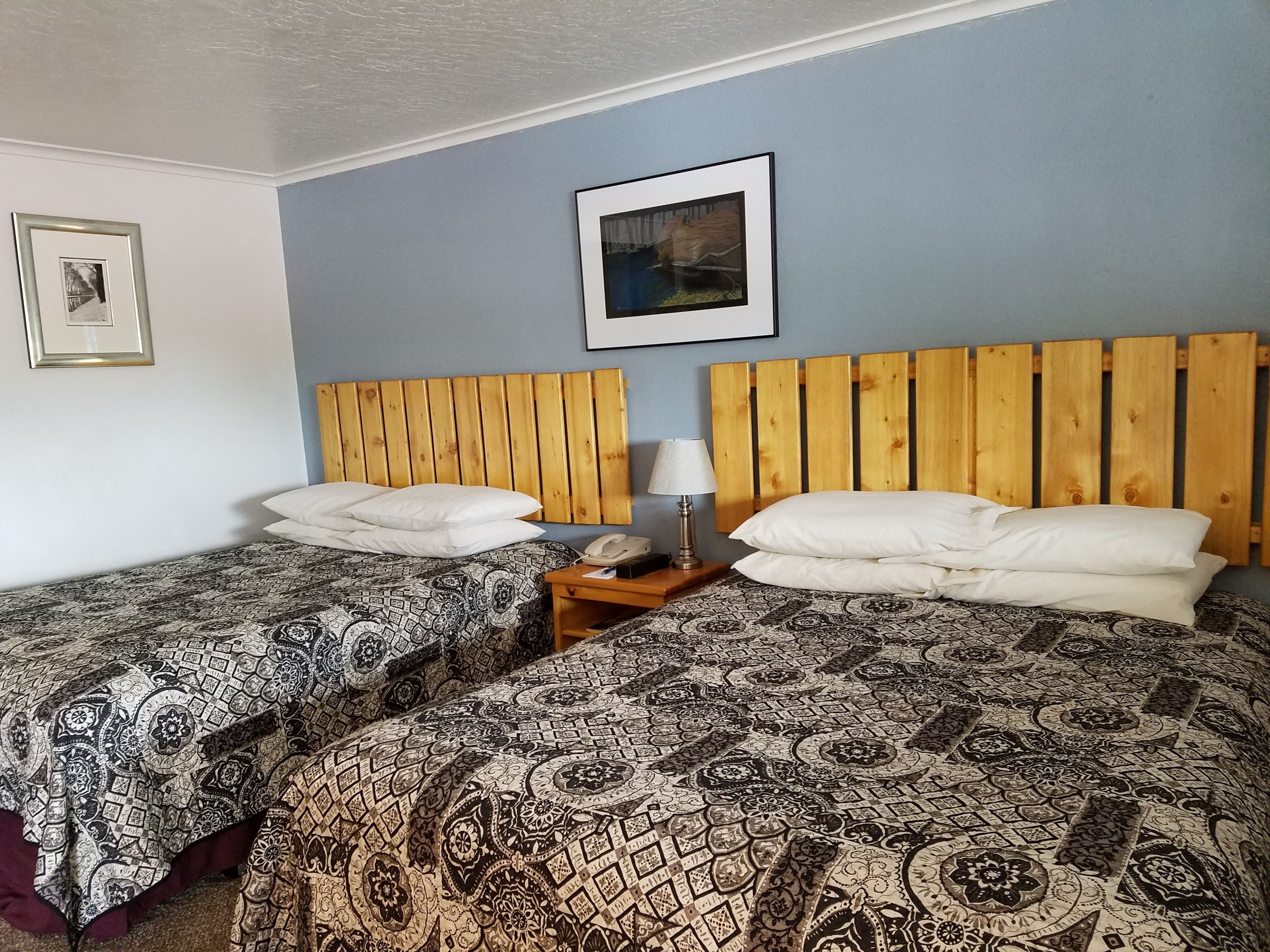 Quarto com duas camas, cada uma com roupa de cama estampada e cabeceira de madeira. Há um quadro na parede e uma mesa de cabeceira com uma luminária.