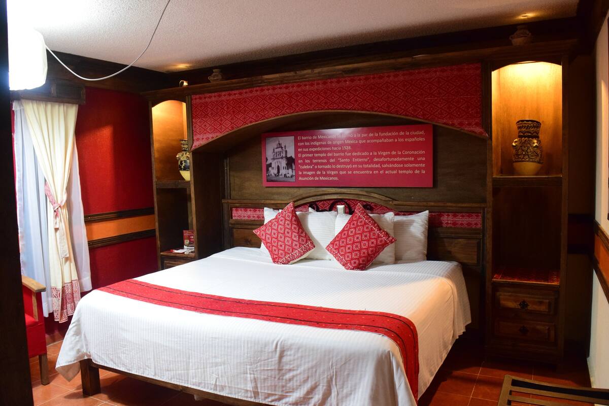 Uma cama bem arrumada com um cobre-leito vermelho e branco, uma cabeceira de madeira e uma mesinha de cabeceira com uma luminária.