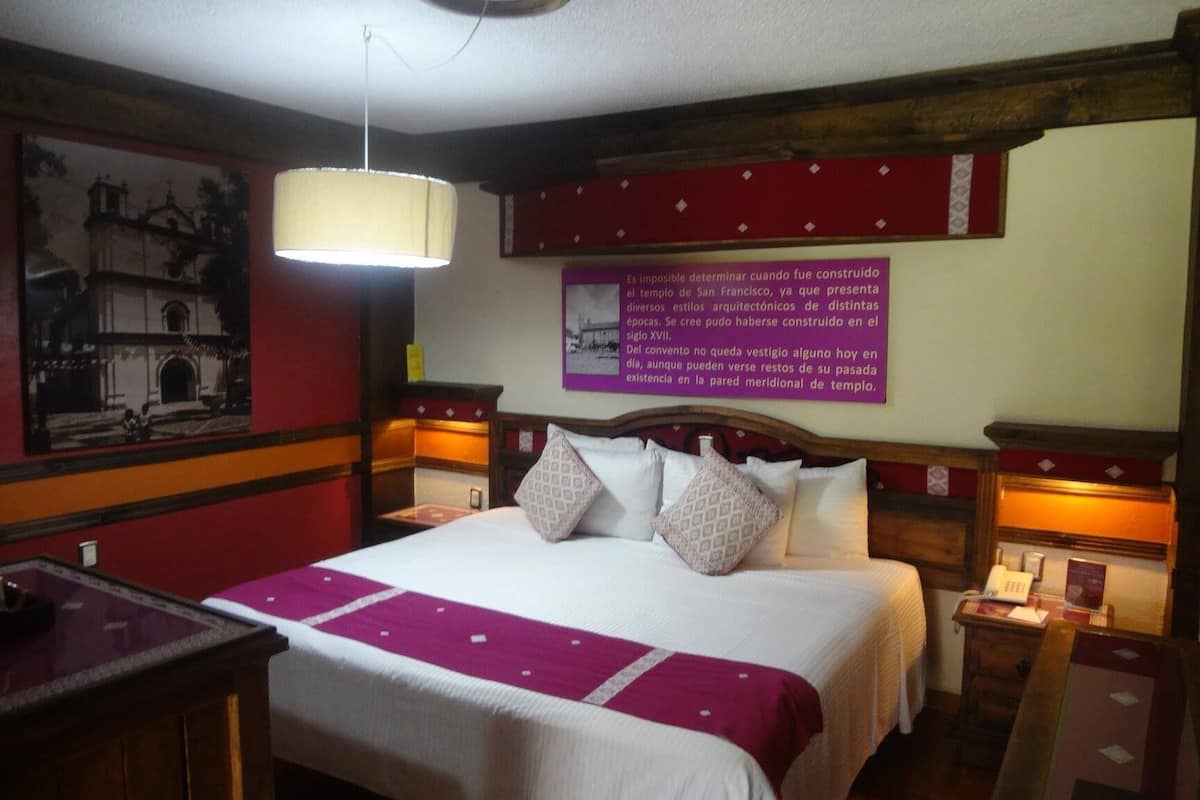 Quarto de hotel com uma cama grande, uma escrivaninha, uma luminária e uma parede com uma fotografia e texto históricos.