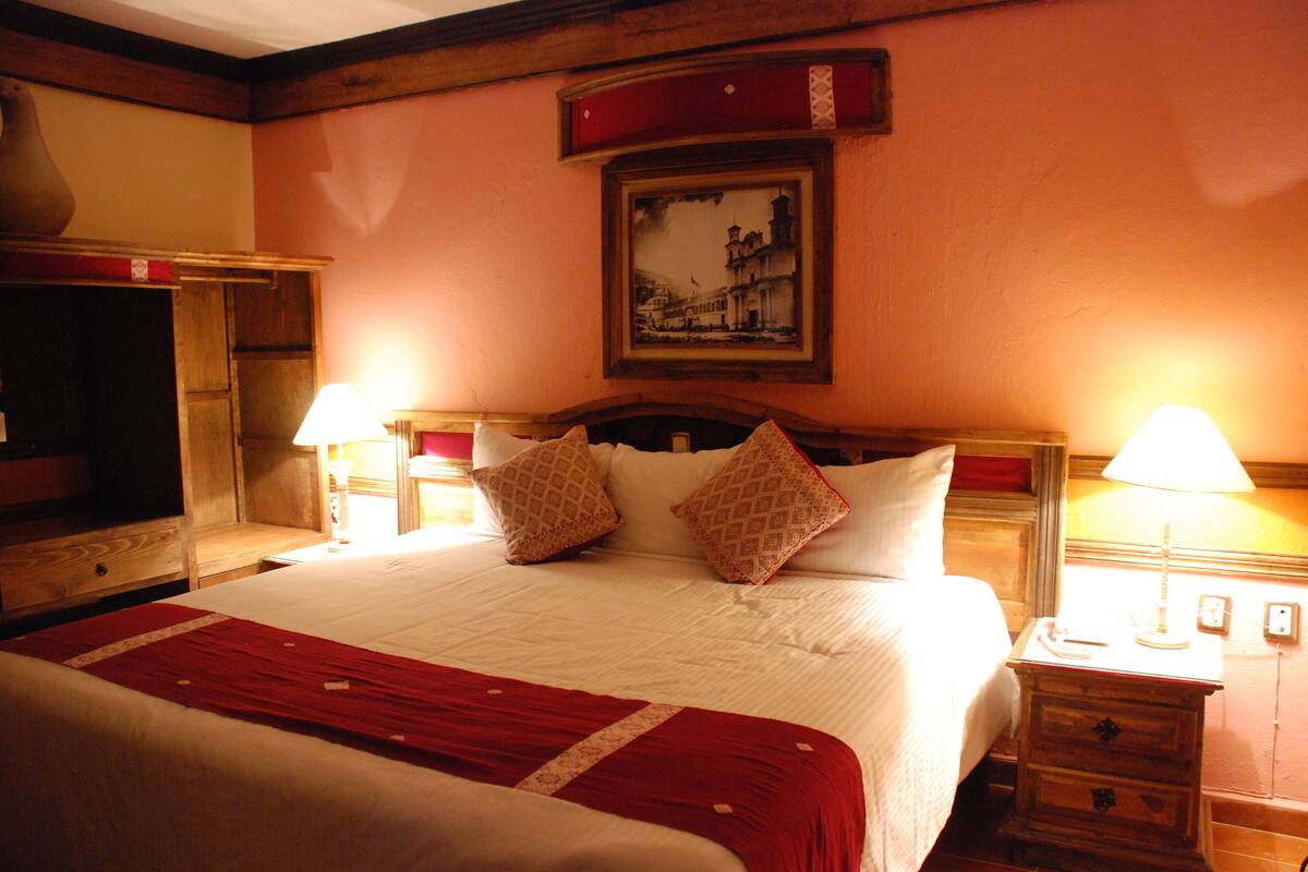 Uma cama bem arrumada com colcha vermelha, duas almofadas decorativas e duas luminárias de cabeceira.