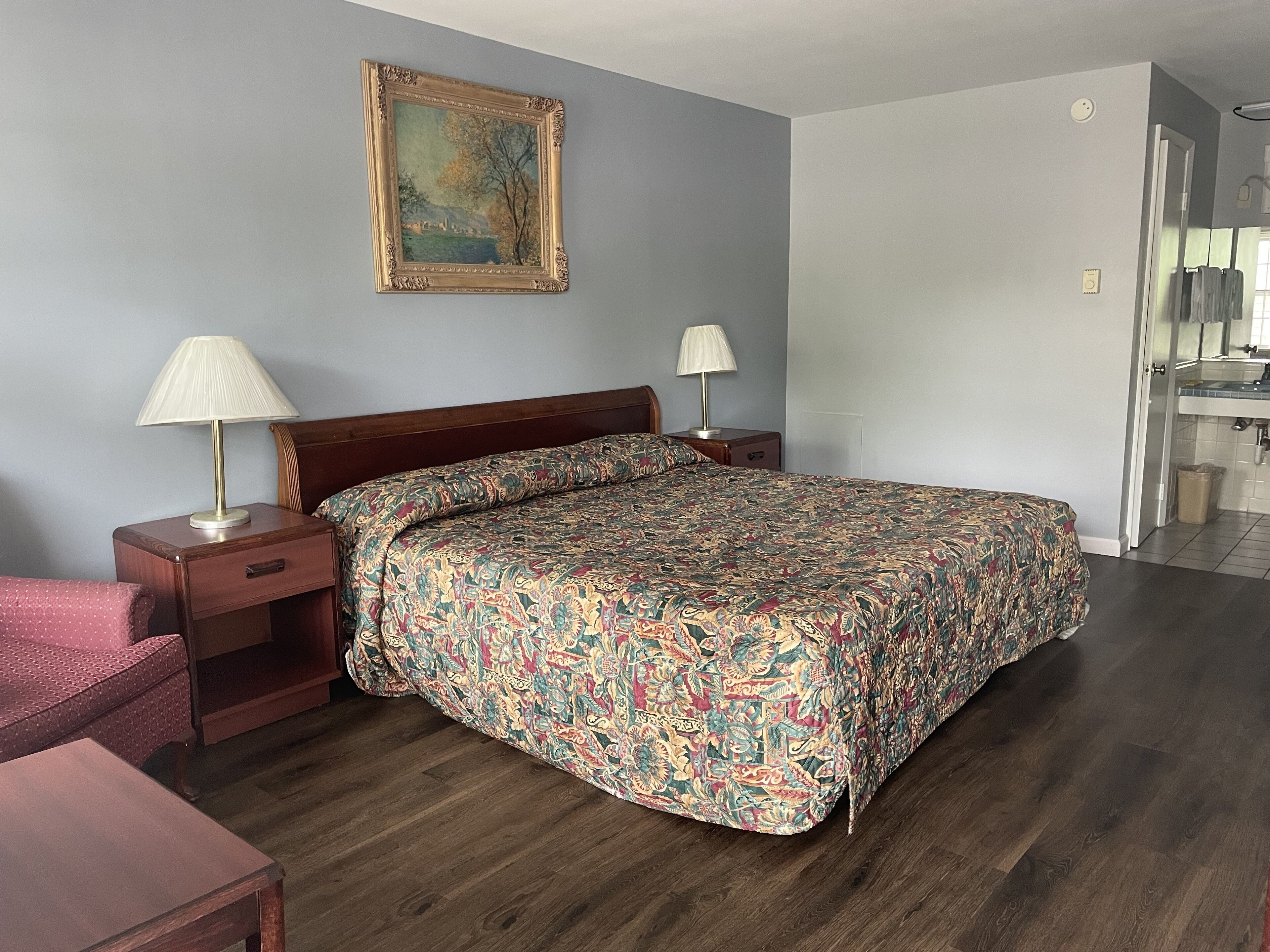 Quarto com cama de madeira, duas mesas de cabeceira com luminárias, uma cadeira, um quadro na parede e um banheiro visível através de uma porta aberta.