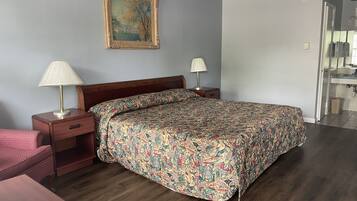 Quarto com cama de madeira, duas mesas de cabeceira com luminárias, uma cadeira, um quadro na parede e um banheiro visível através de uma porta aberta.