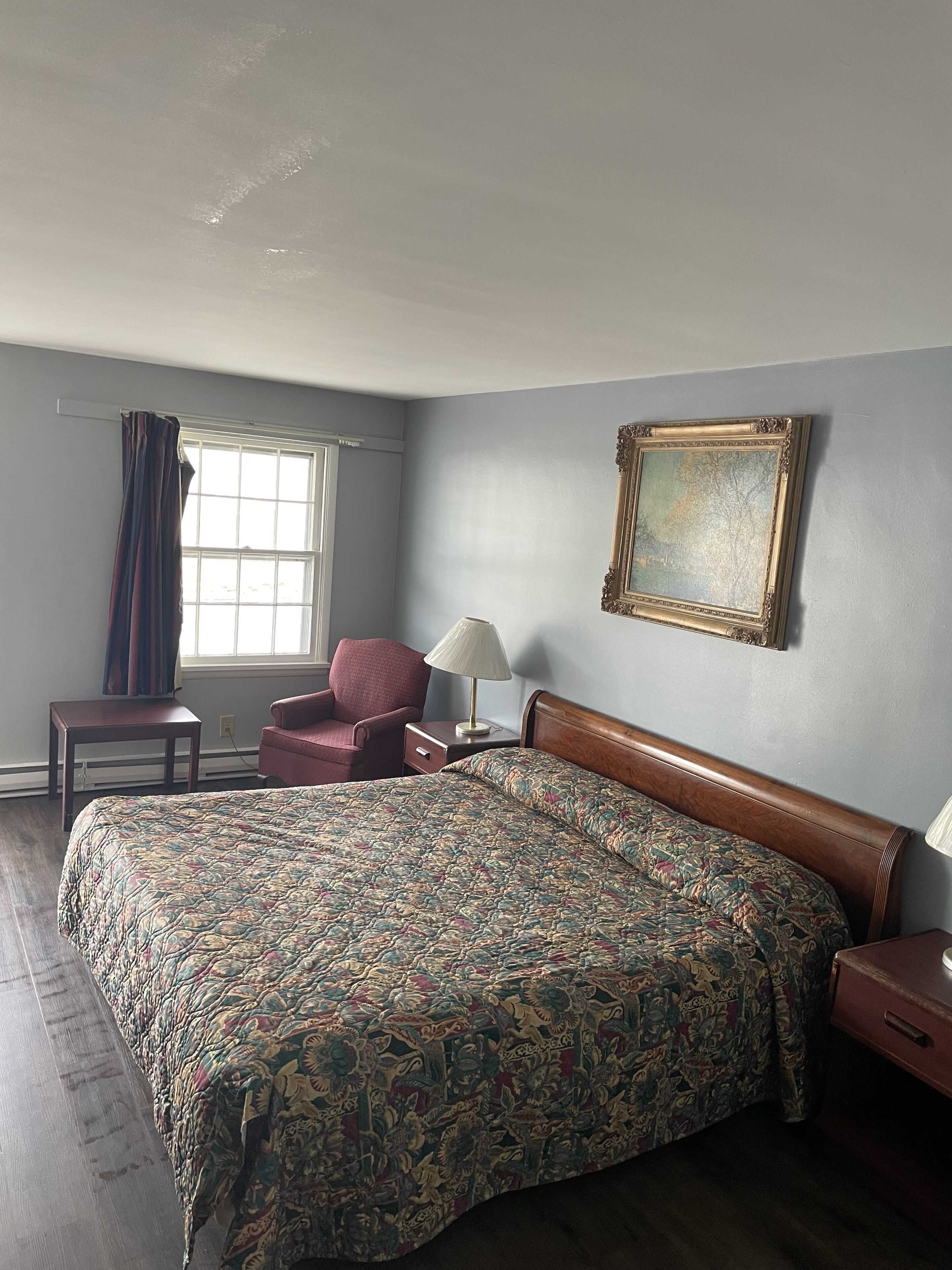Um quarto com uma cama grande, uma cadeira, uma mesa, um abajur e um quadro na parede.