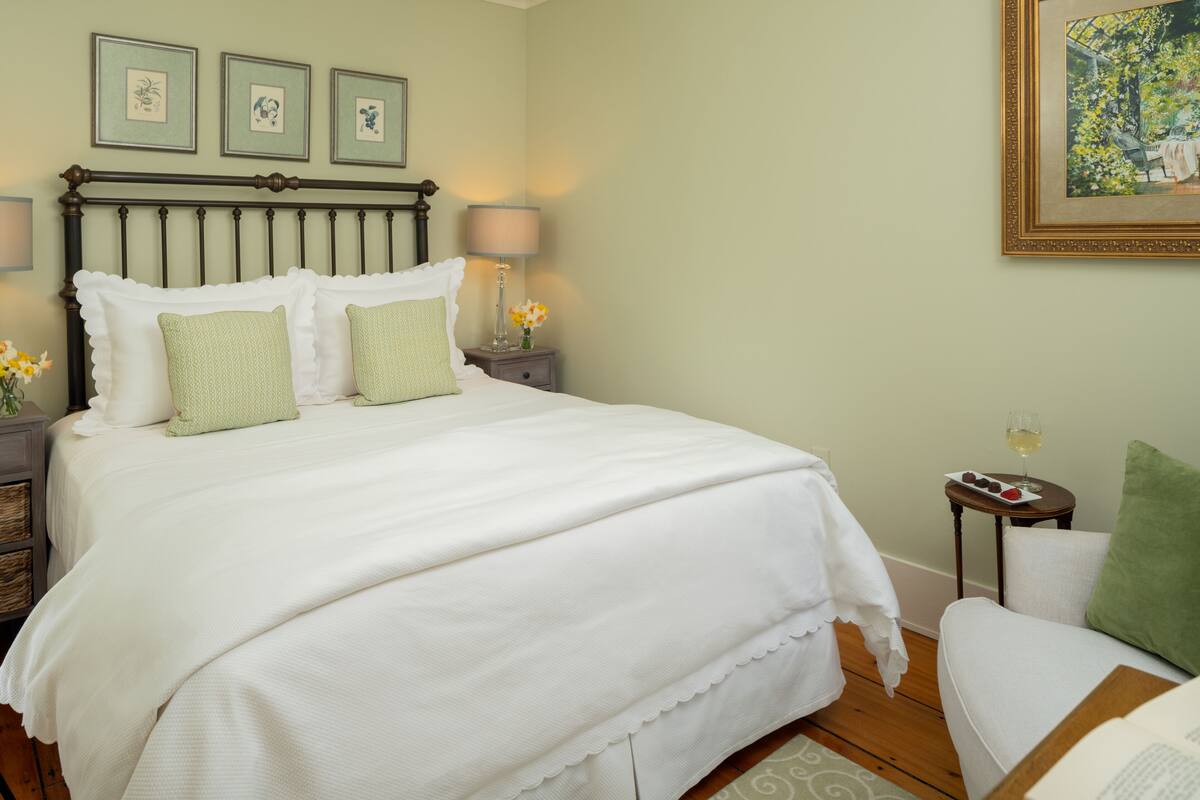 Cama bem arrumada com roupa de cama branca e almofadas verdes, mesa de cabeceira com abajur e quadro na parede.