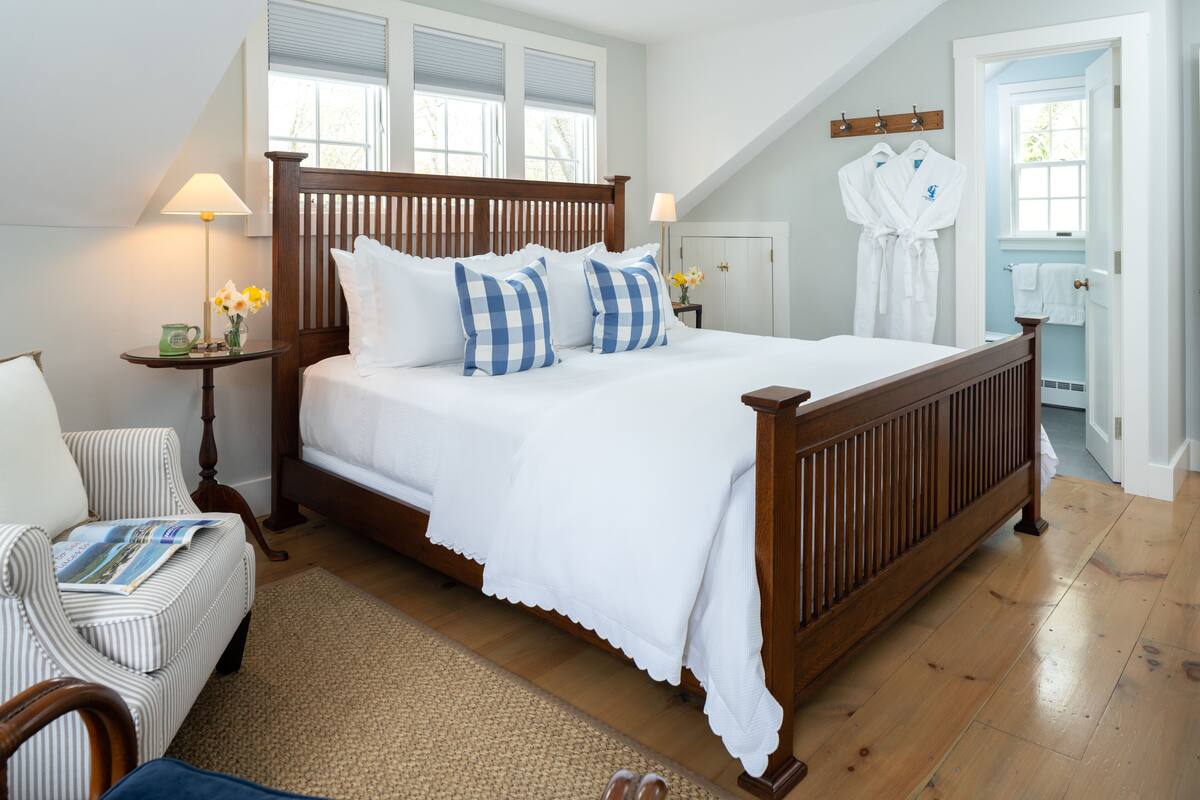 Quarto com cama de madeira, duas mesas de cabeceira com luminárias, uma cadeira e vista para um banheiro com toalhas e roupões.