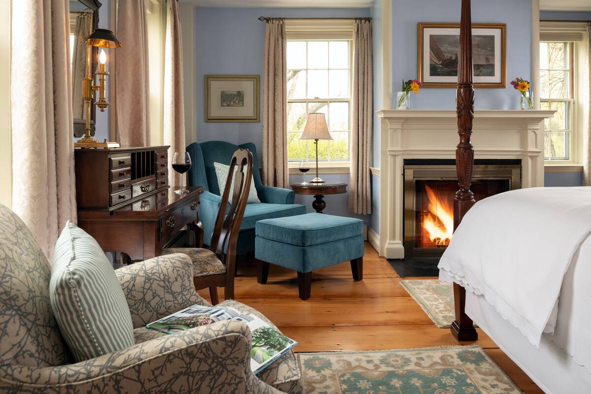 Quarto com uma cama grande, uma cadeira de madeira, um banquinho azul, cômoda, luminária e lareira.