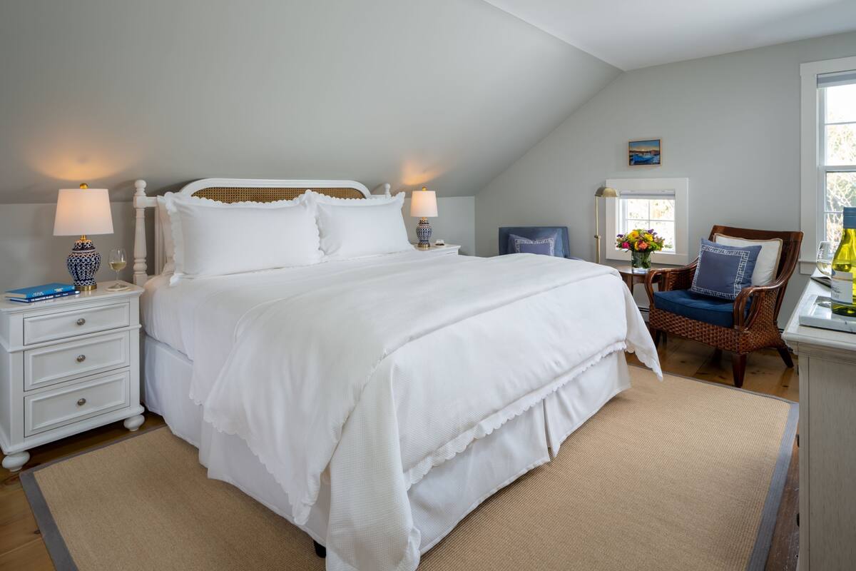 Quarto com cama grande, mesa de cabeceira, cadeira de vime e escrivaninha com abajur.