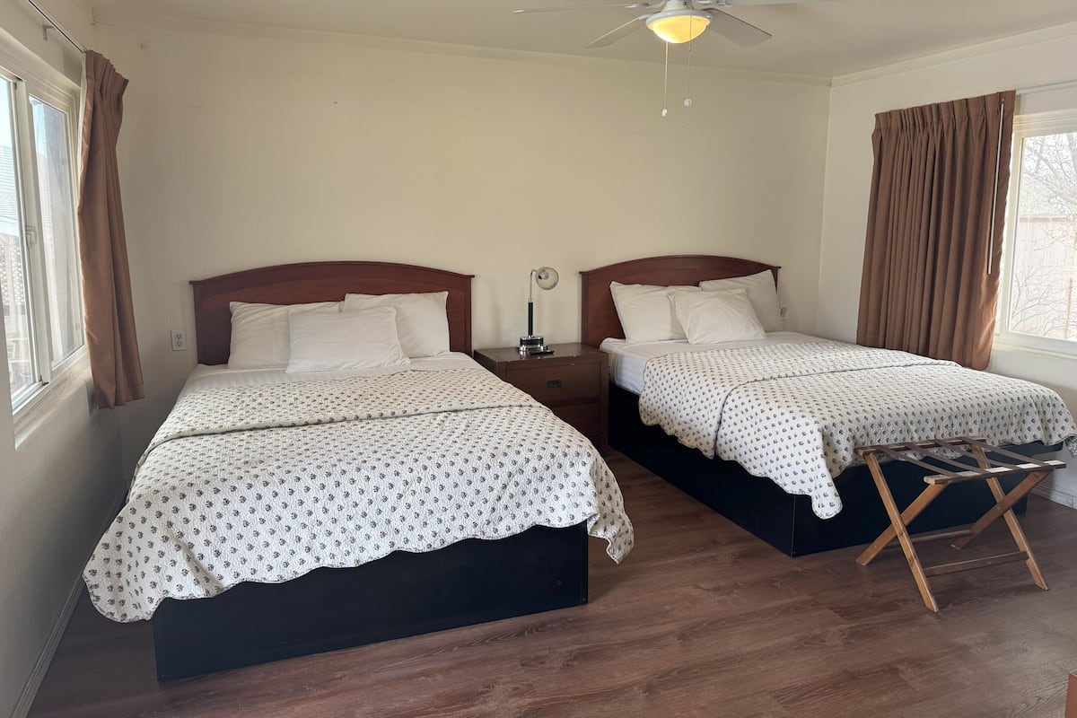 Quarto com duas camas, um ventilador de teto, uma mesinha de cabeceira e uma janela com cortinas.