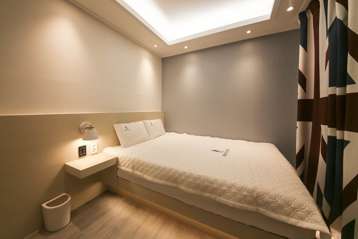 Quarto moderno com claraboia, mesa de cabeceira com abajur, cama com edredom branco e cortina estampada.