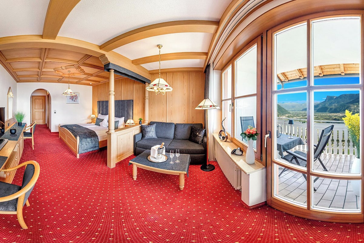 Um quarto espaçoso com carpete vermelho, uma janela grande com vista, um sofá, uma mesa de jantar e uma cama.