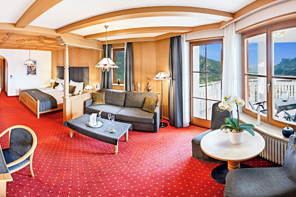 Quarto de hotel com carpete vermelho, um sofá, uma mesa de centro, área de jantar com mesa e cadeiras, e grandes janelas com vista para as montanhas.