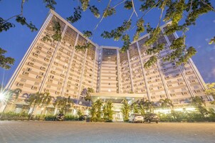 Um hotel alto com vários andares, entrada central e vegetação ao redor.