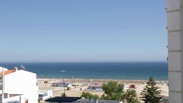 Vista da praia a partir de uma varanda, com céu claro, areal extenso e alguns edifícios ao primeiro plano.