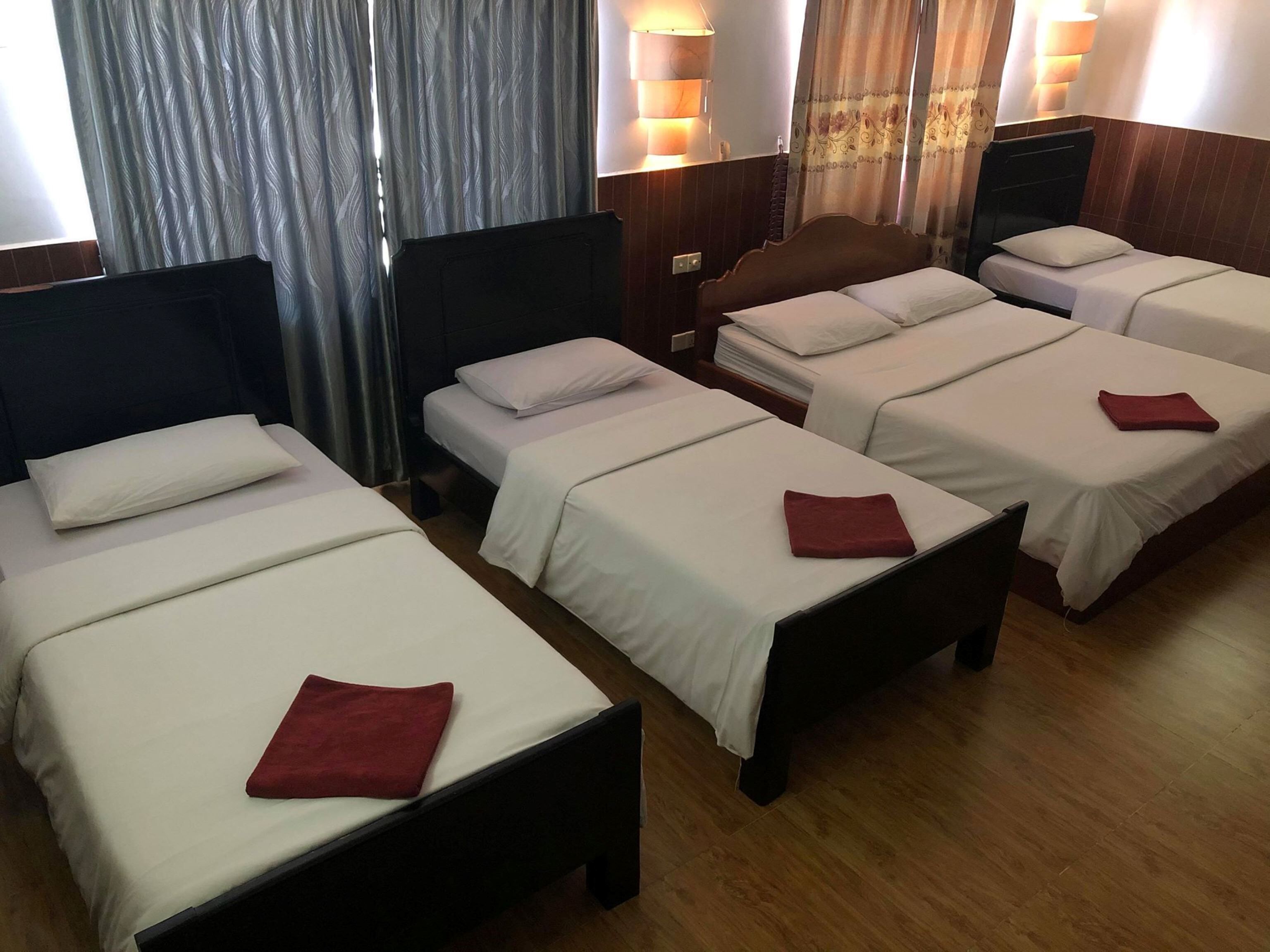 Quarto de hotel com quatro camas individuais, cada uma com roupa de cama branca e toalha vermelha. O quarto possui piso de madeira, cabeceira de madeira e uma janela com cortinas.