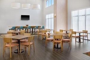 Uma sala de jantar ampla com várias mesas e cadeiras, uma TV de tela plana na parede e grandes janelas que proporcionam luz natural.