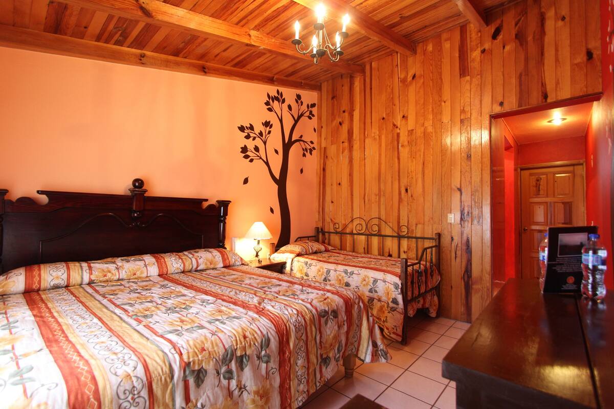 Quarto de madeira com duas camas, um candelabro e uma parede de madeira decorada com um adesivo de árvore.