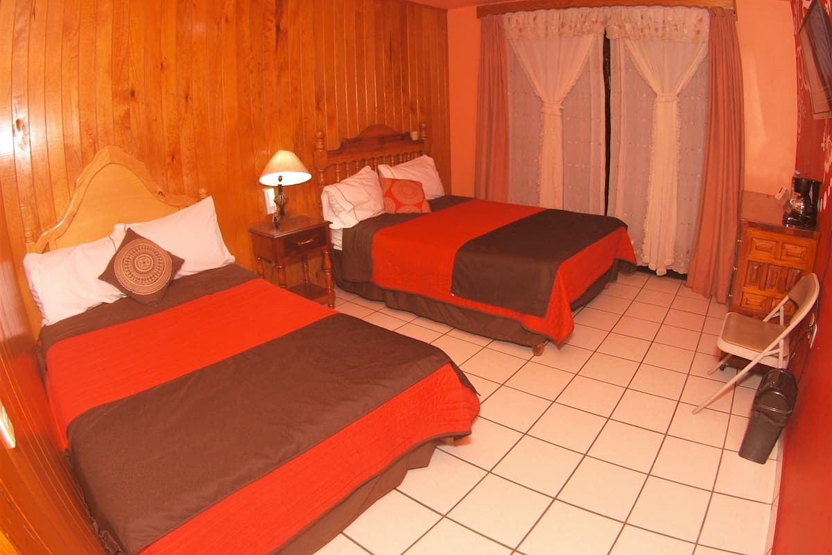 Quarto de hotel com duas camas, paredes de madeira, piso ladrilhado, mesa de cabeceira com luminária, uma cadeira e uma janela com cortinas.
