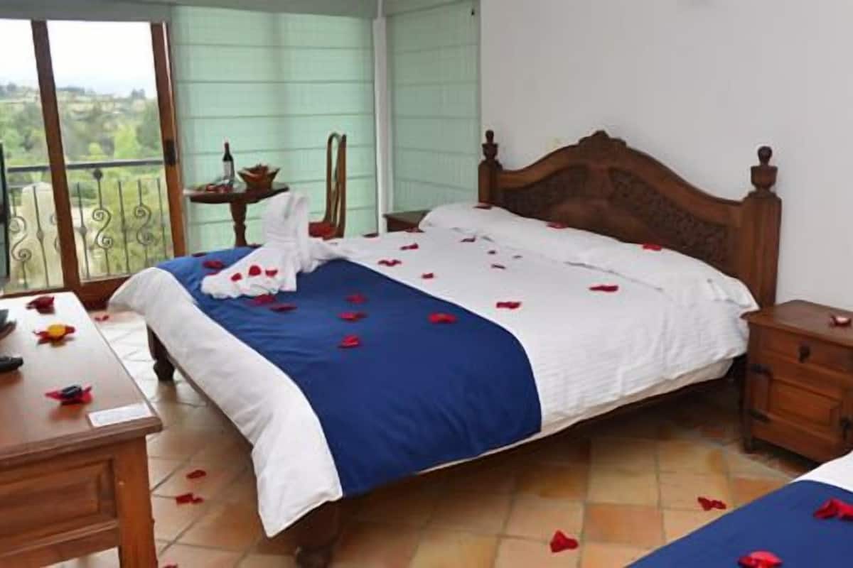 Quarto de hotel com cama, mesa de cabeceira de madeira, escrivaninha com cadeira e janela com vista para vegetação.