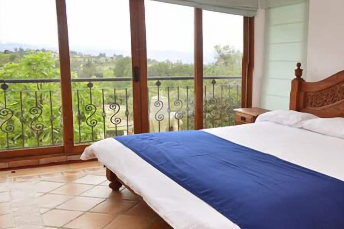 Quarto com cama grande, cabeceira de madeira, mesa de cabeceira e vista para o verde através de uma varanda.