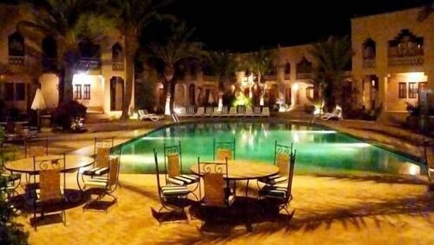 Vista noturna de uma área de piscina de hotel com espreguiçadeiras e mesas, cercada por palmeiras e edifícios iluminados.