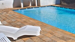 Uma piscina retangular com água azul cristalina, rodeada por um deck ladrilhado e espreguiçadeiras brancas.