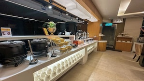 Disposição de buffet com diversos recipientes de comida, uma estação de café e uma placa indicando a localização.