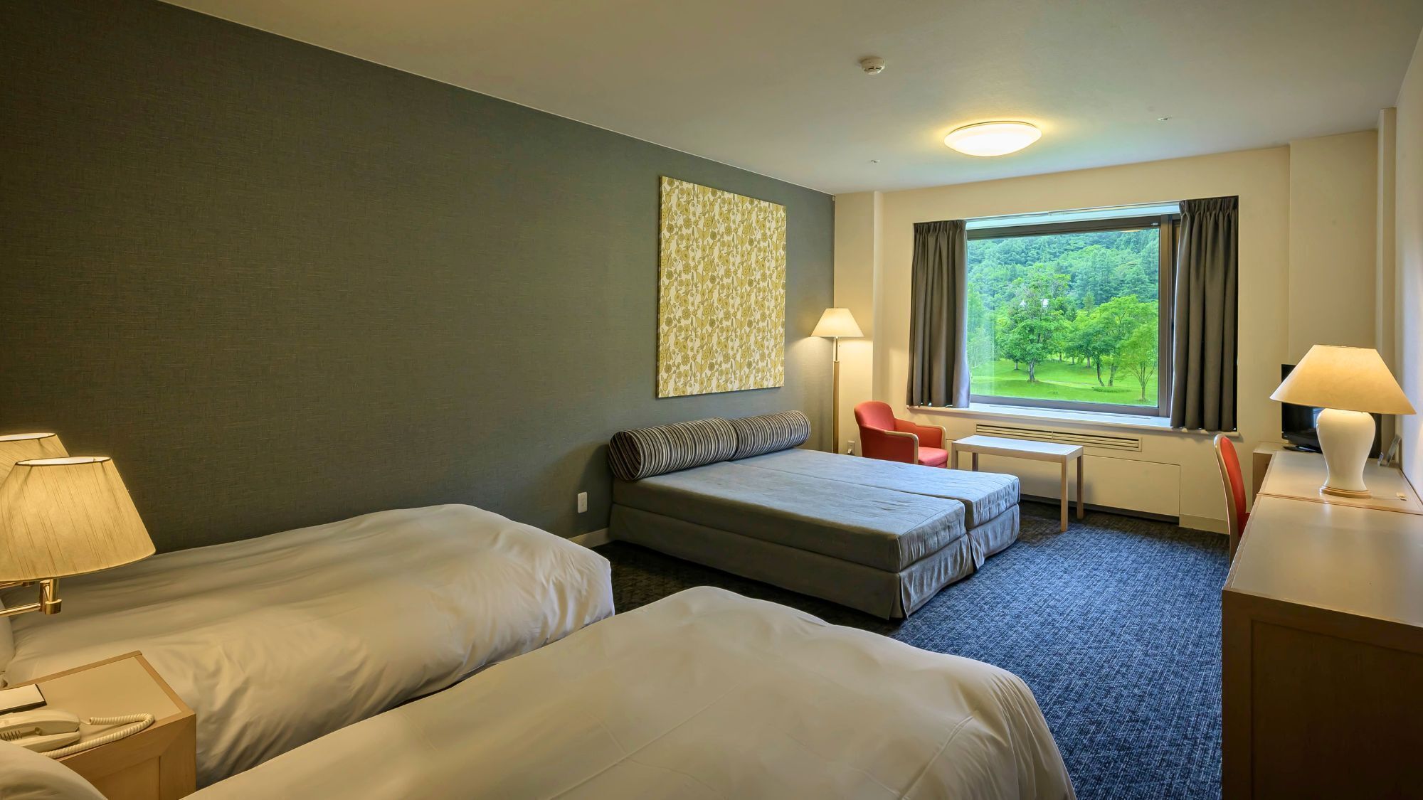 Quarto de hotel com duas camas, uma escrivaninha, uma cadeira e uma janela com vista para o verde.