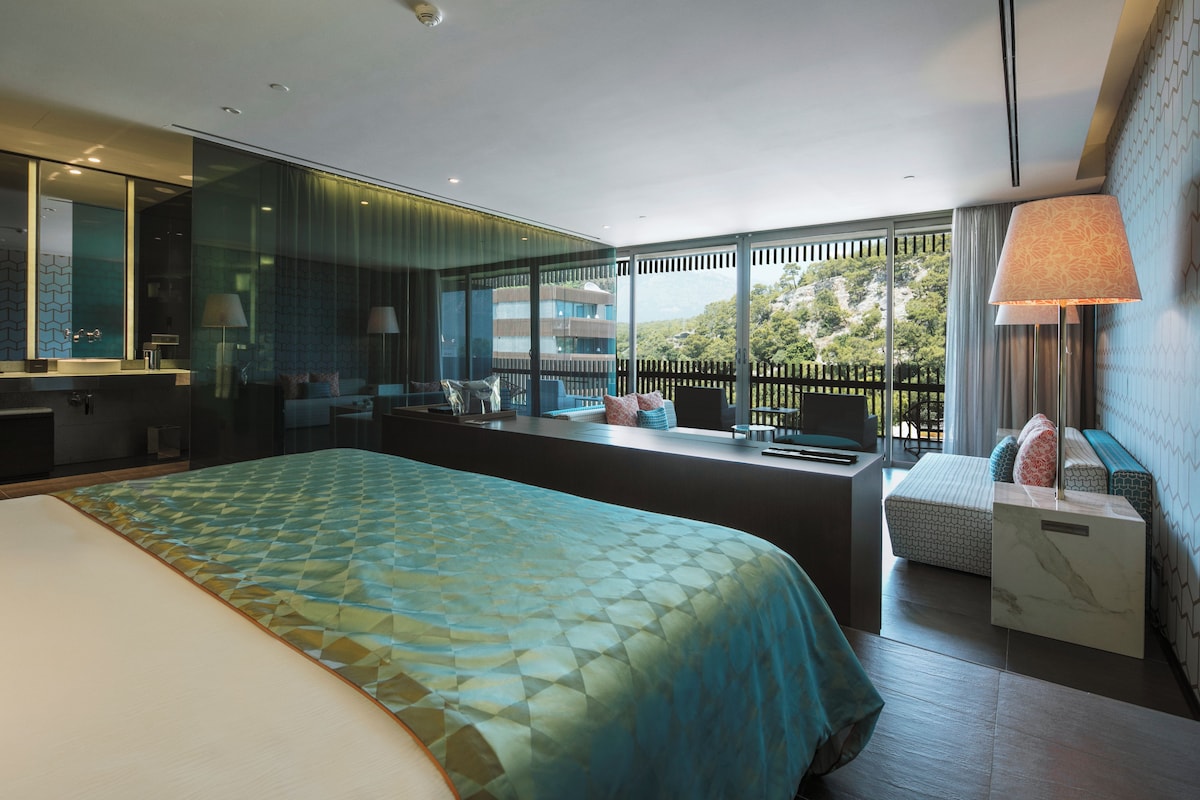 Quarto de hotel moderno com uma cama grande, área de estar com vista, banheiro com divisória de vidro e varanda com vista.