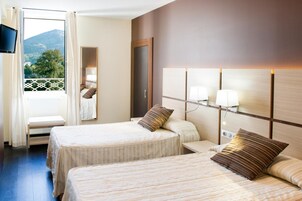 Quarto de hotel com duas camas, varanda com vista para uma montanha, televisão e mesa de cabeceira com luminárias.