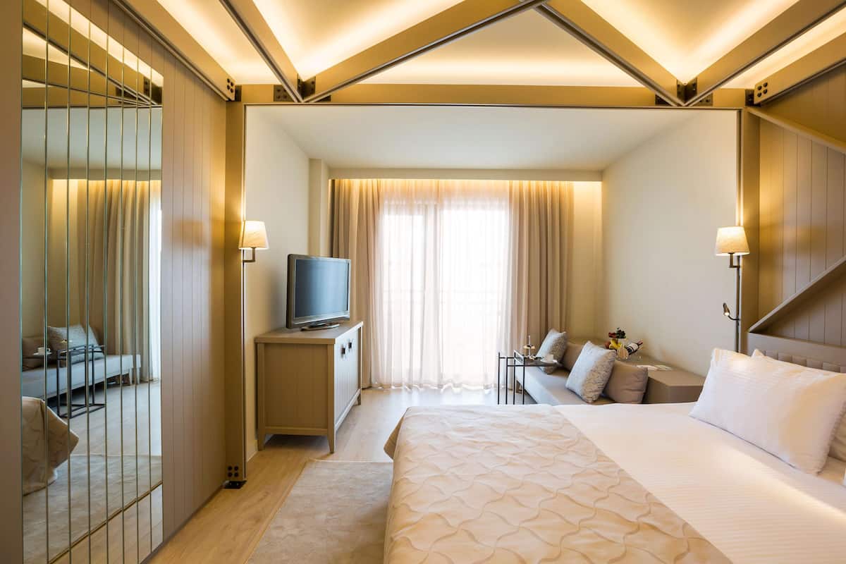 Quarto de hotel com uma cama grande, uma televisão sobre uma mesa de cabeceira de madeira, uma escrivaninha com abajur e vista para o exterior através de cortinas leves.