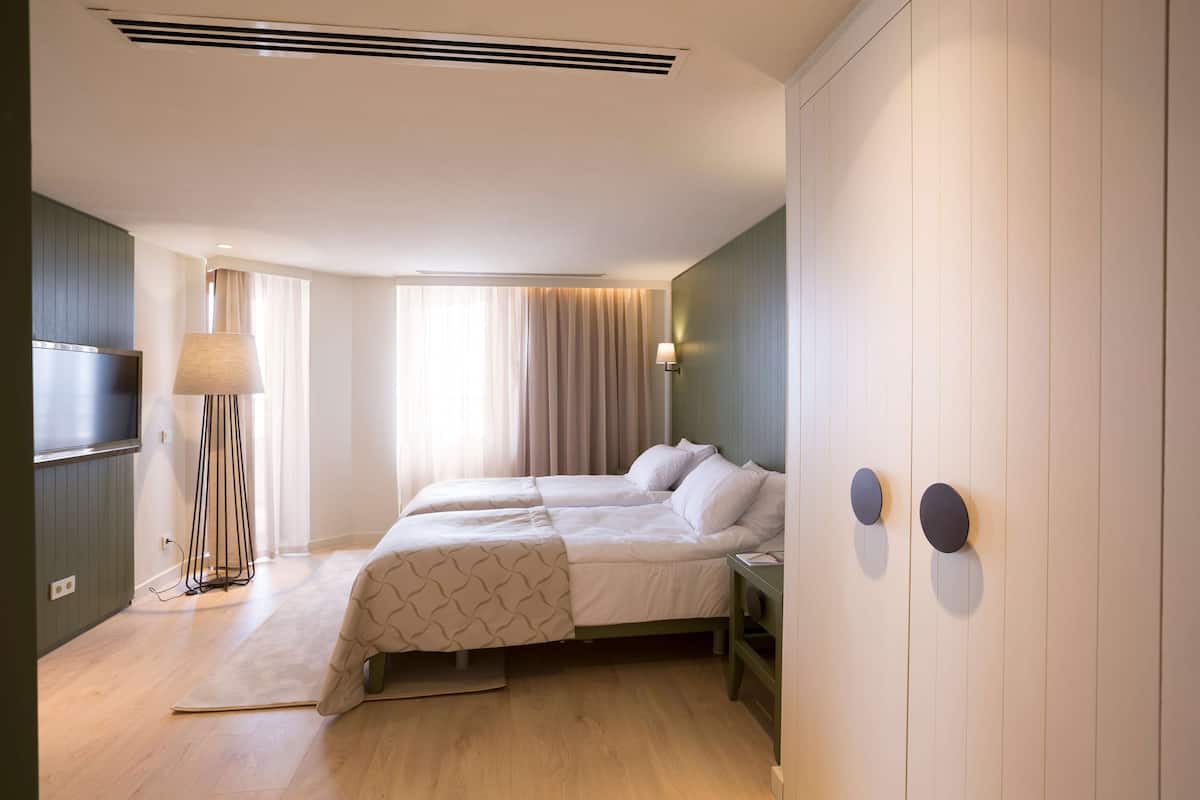 Quarto de hotel com duas camas, televisão, candeeiro de chão e uma janela grande com cortinas.