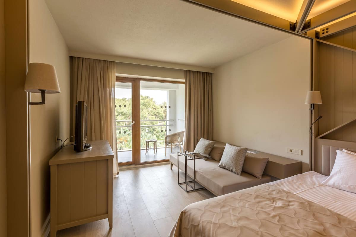 Quarto de hotel com cama, sofá, escrivaninha com televisão e varanda com cortinas.