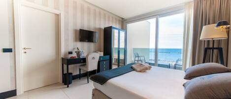 Quarto de hotel com cama, escrivaninha, cadeira, televisão, guarda-roupa e vista para o mar.