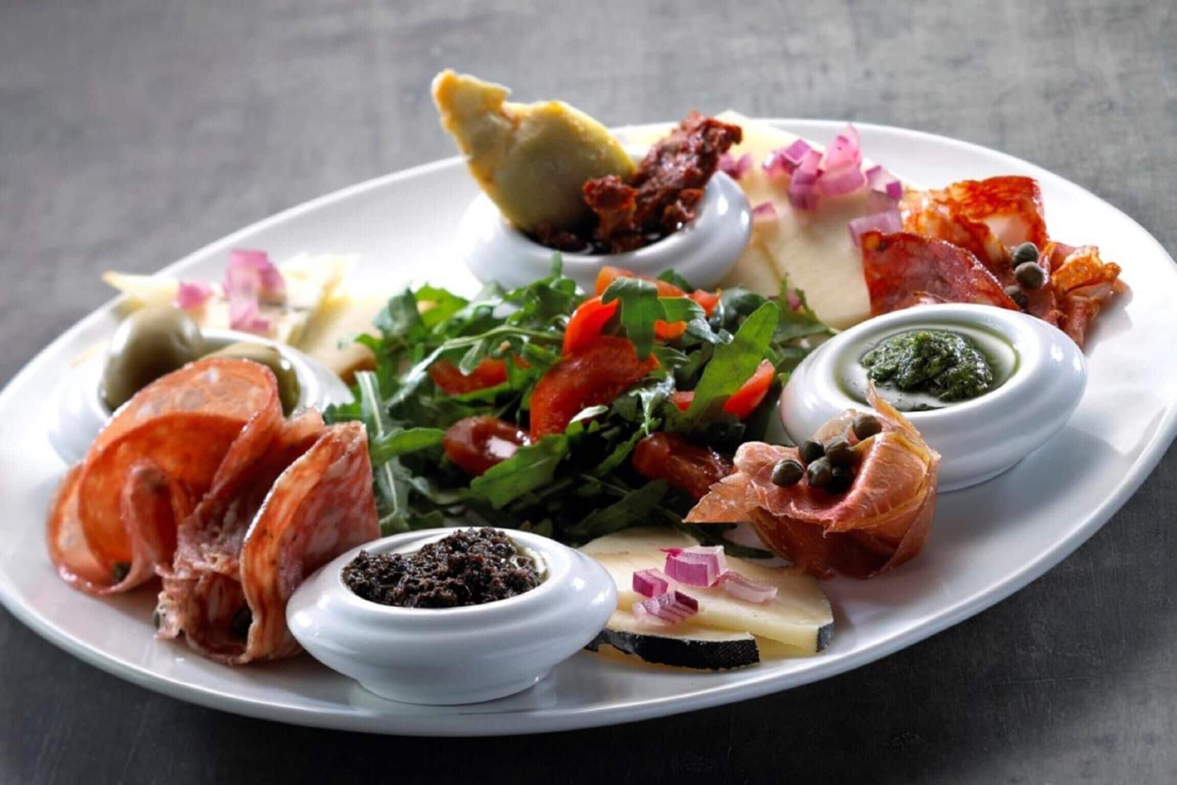 Um prato com diversos petiscos, incluindo embutidos, queijos e uma salada com acompanhamentos variados.