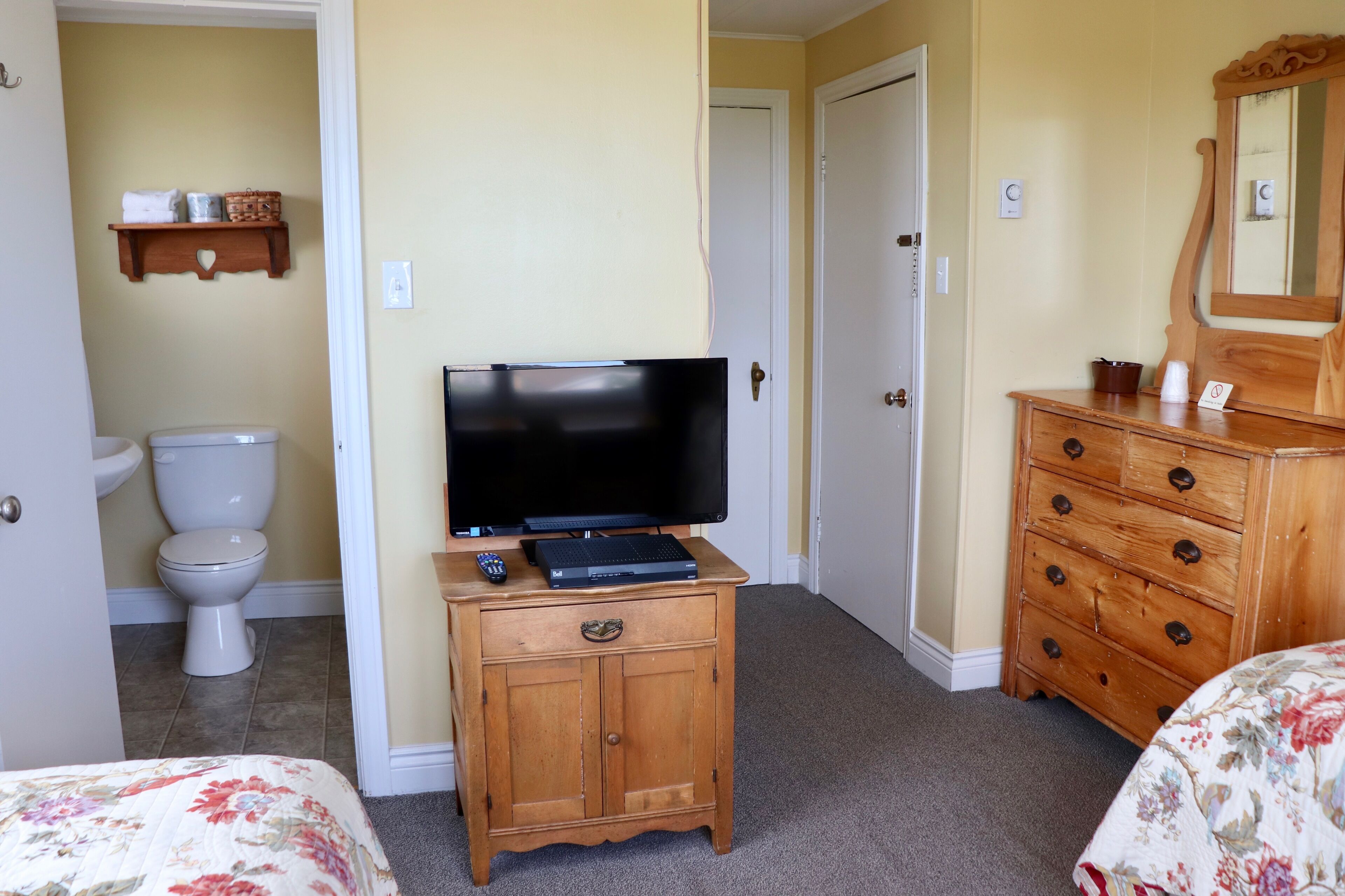 Quarto com cômoda de madeira, televisão sobre um suporte, cama com roupa de cama floral, mesa de cabeceira de madeira e banheiro com vaso sanitário e prateleira.