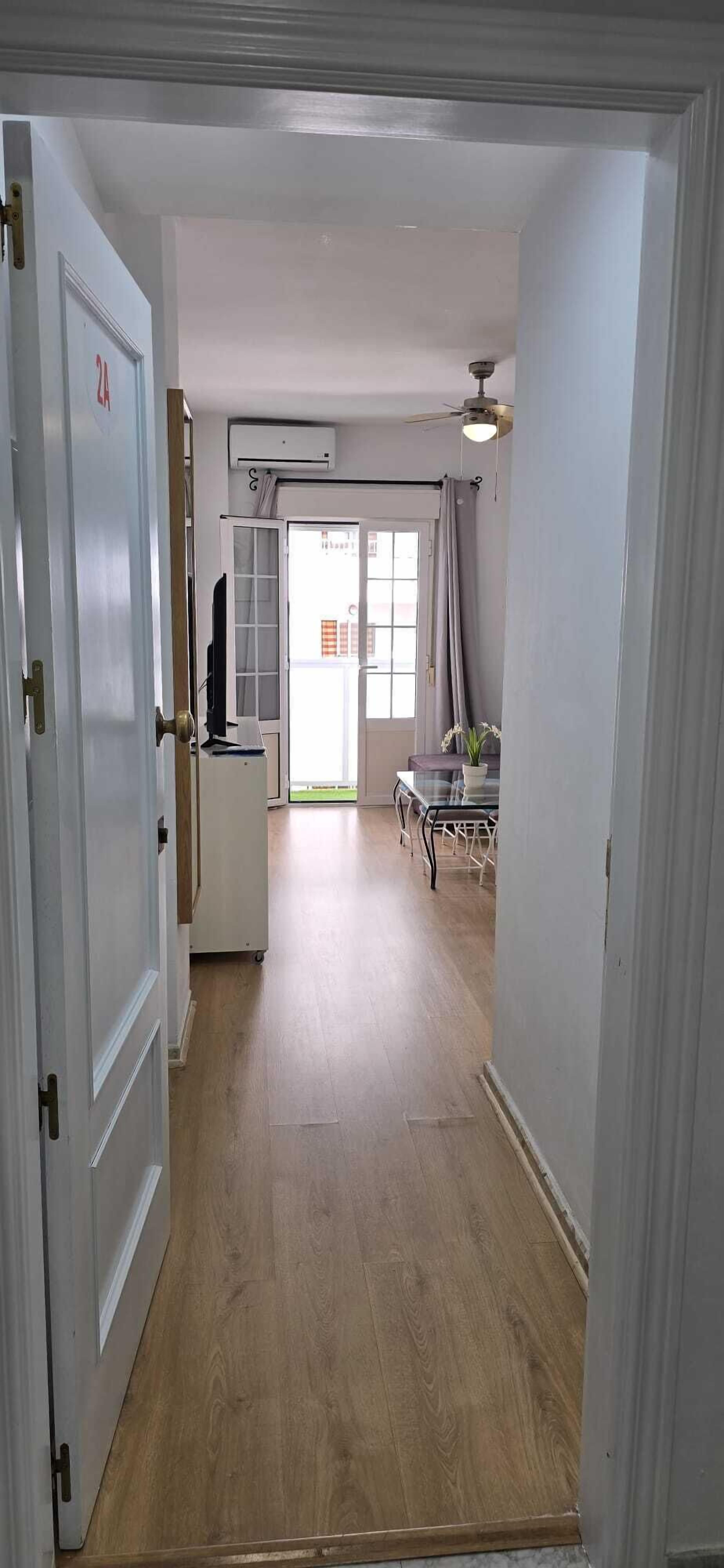 Um corredor com piso de madeira que leva a um quarto com ventilador de teto e janela com cortinas.