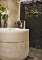 Uma área de recepção com um balcão moderno, um vaso com flores brancas e uma parede com o texto "LE SPA by SOTHYS PARIS".