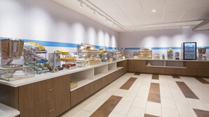 Uma área de buffet com diversos pontos de serviço, incluindo uma exposição de frutas e um dispensador de bebidas.