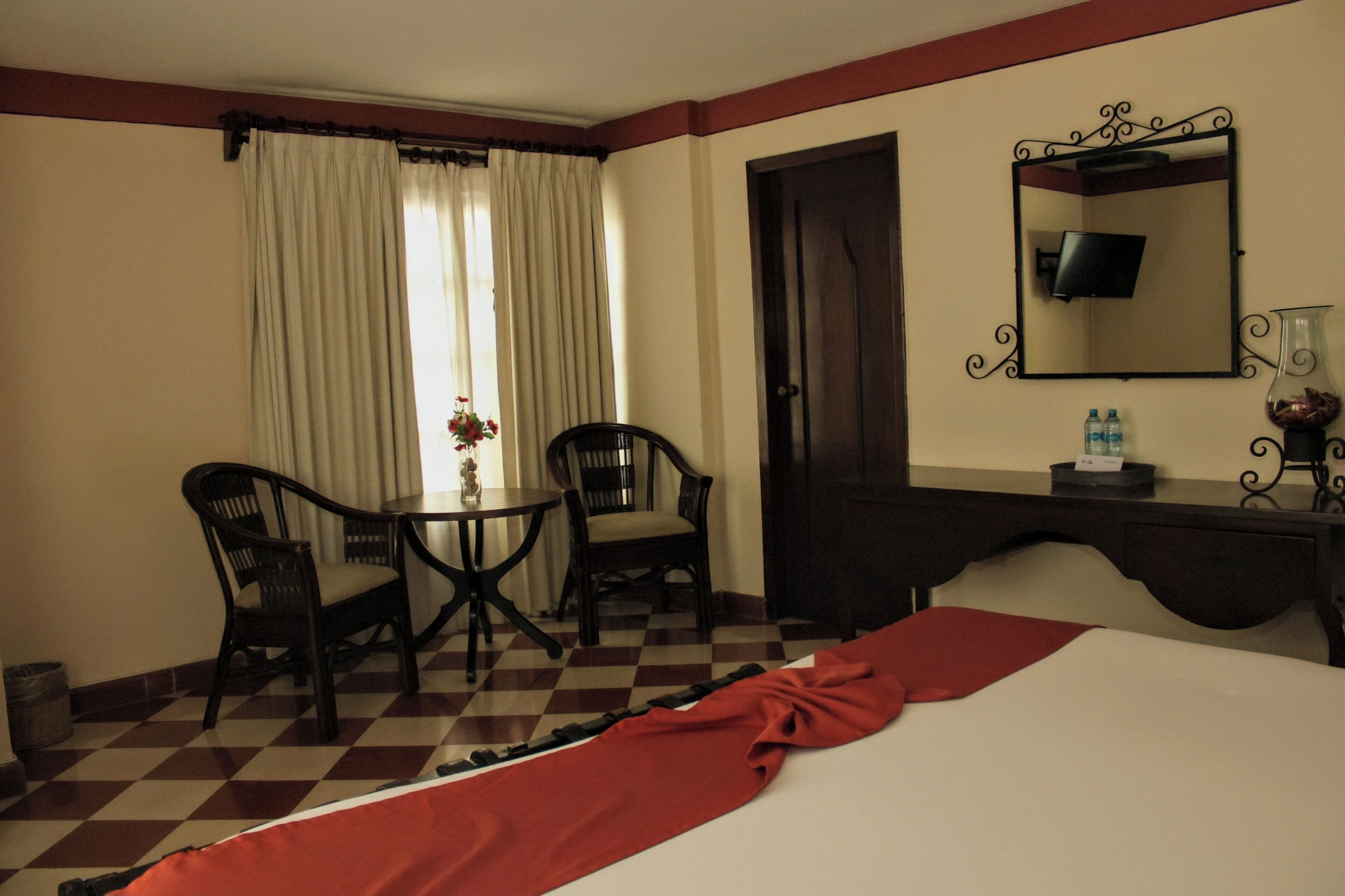 Quarto de hotel com piso xadrez, cama com um corredor vermelho, duas cadeiras, uma pequena mesa redonda com um vaso, um espelho e uma porta.