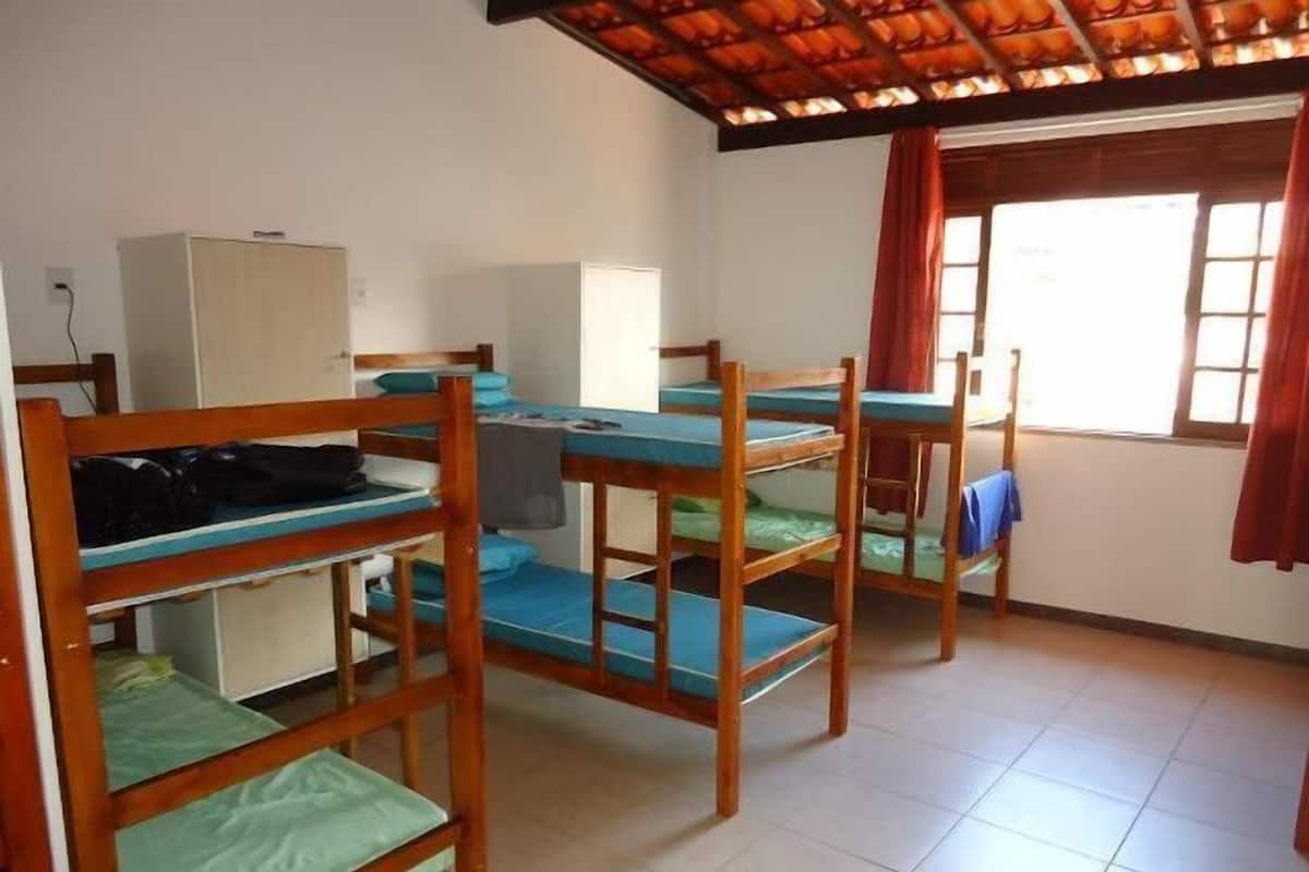 Quarto em dormitório com beliches, uma escrivaninha e janela com cortinas.