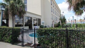 Um hotel com piscina, cercado por uma cerca de ferro preto e palmeiras.