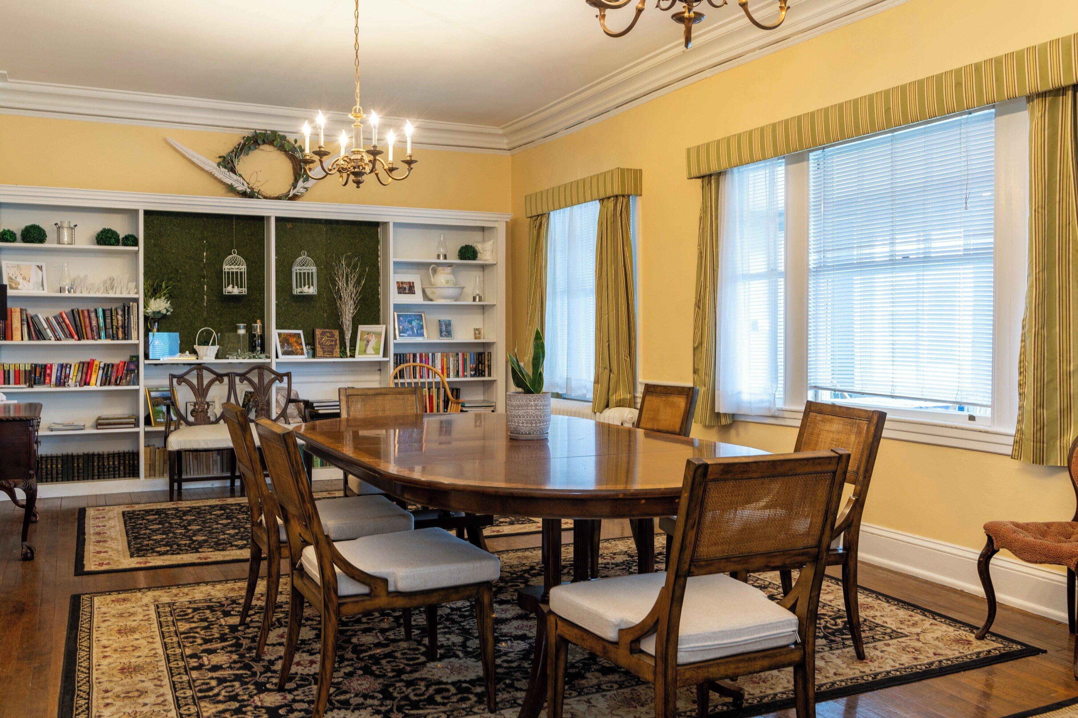 Uma sala de jantar com uma grande mesa de madeira, seis cadeiras e uma estante repleta de livros e objetos decorativos.